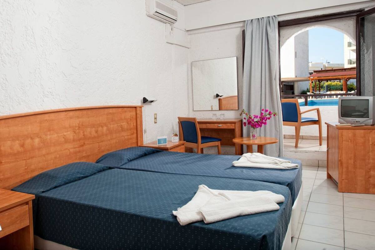 Quarto de hotel com cama de madeira, escrivaninha, cadeira, televisão e vista para a piscina.