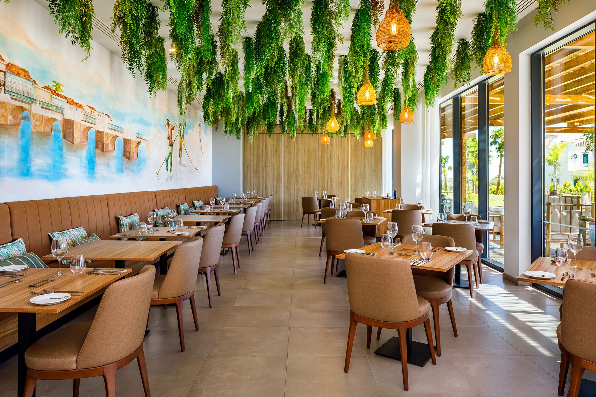 Um restaurante com mesas e cadeiras de madeira, um mural na parede, plantas pendentes e grandes janelas.