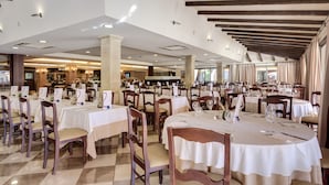 Uma ampla sala de jantar com várias mesas redondas, cadeiras de madeira e toalhas de mesa brancas.