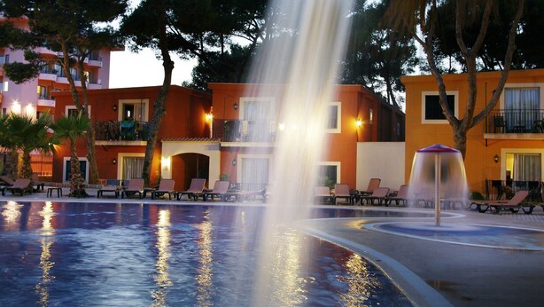Piscina com fonte de água, espreguiçadeiras e um edifício com varandas ao fundo.