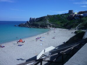 Uma praia com uma escadaria de pedra descendo até a água, um litoral rochoso e edifícios numa encosta.