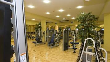 Uma academia com diversos aparelhos de exercícios, piso de madeira e iluminação no teto.
