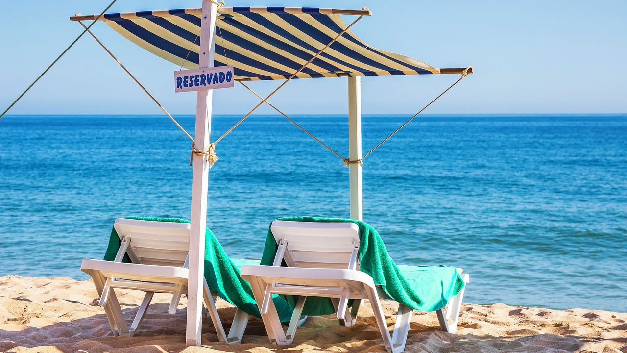 Uma praia com duas cadeiras de praia brancas sob um toldo listrado, uma placa 'RESERVADO' e o oceano ao fundo.