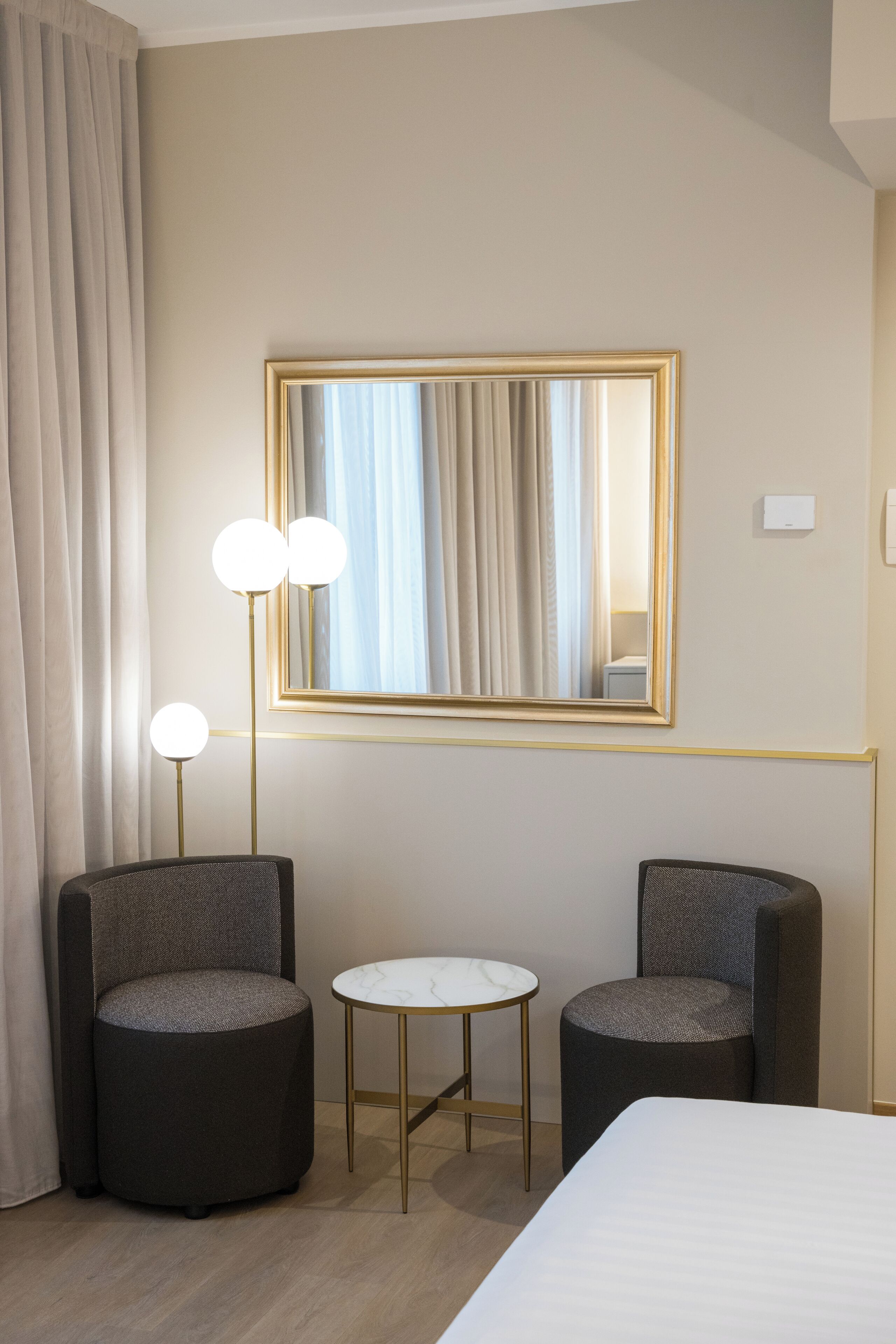 Quarto de hotel com cama, duas poltronas, uma mesinha redonda, abajur e espelho na parede.