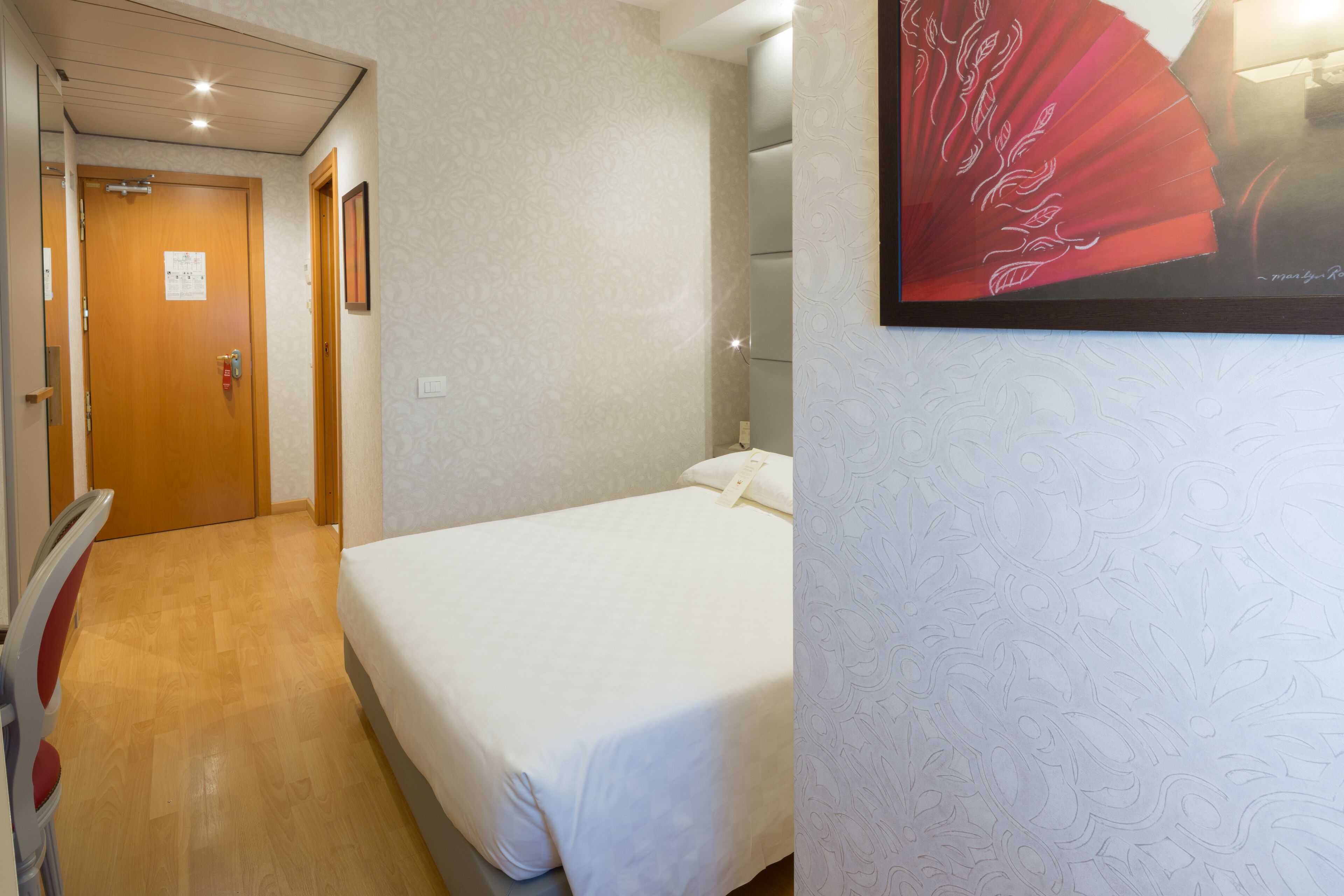 Quarto de hotel com piso de madeira, cama com roupa de cama branca, porta com uma placa e um quadro na parede.