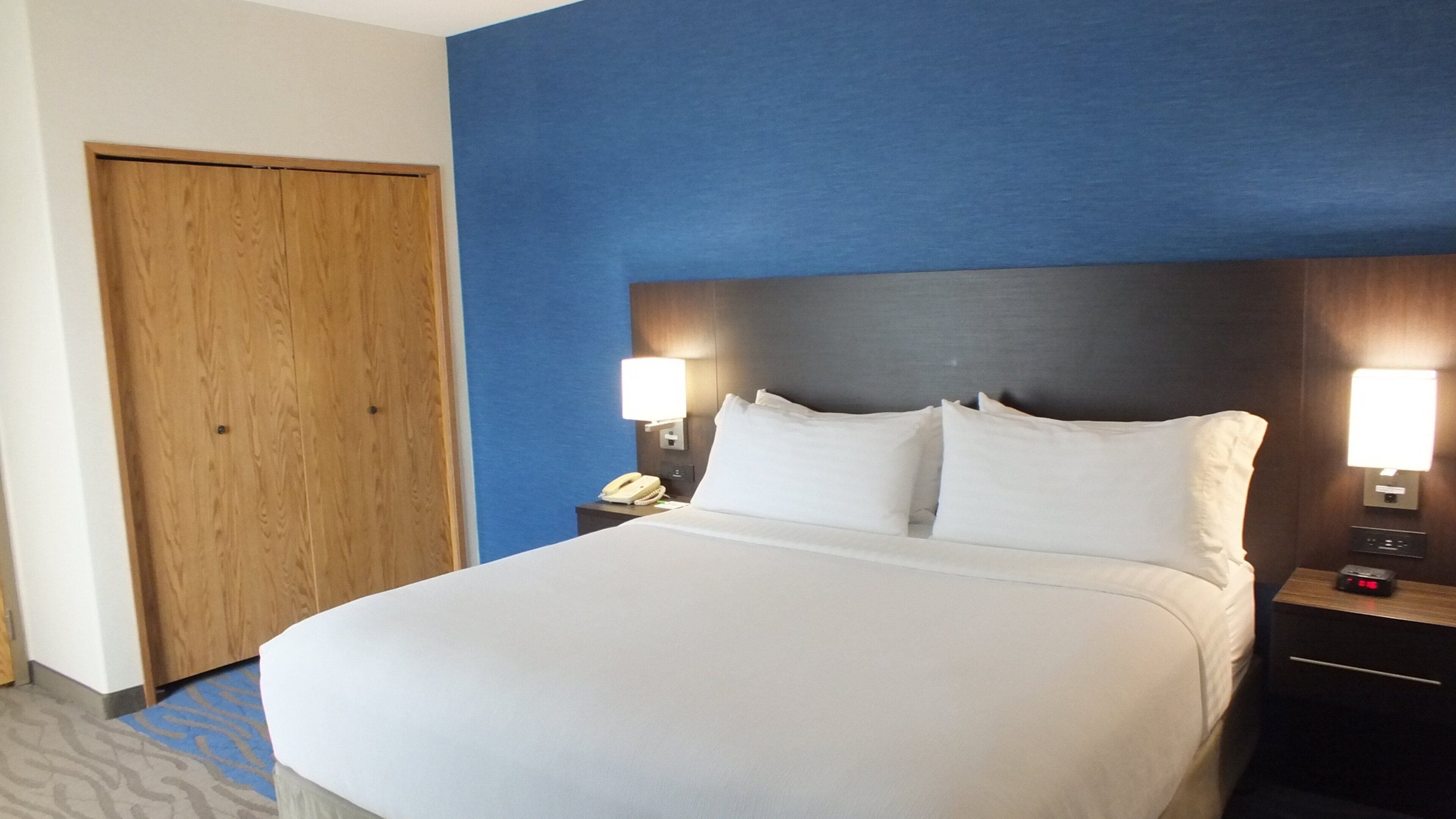 Quarto de hotel com uma cama grande, uma porta de madeira e duas mesas de cabeceira com abajures.