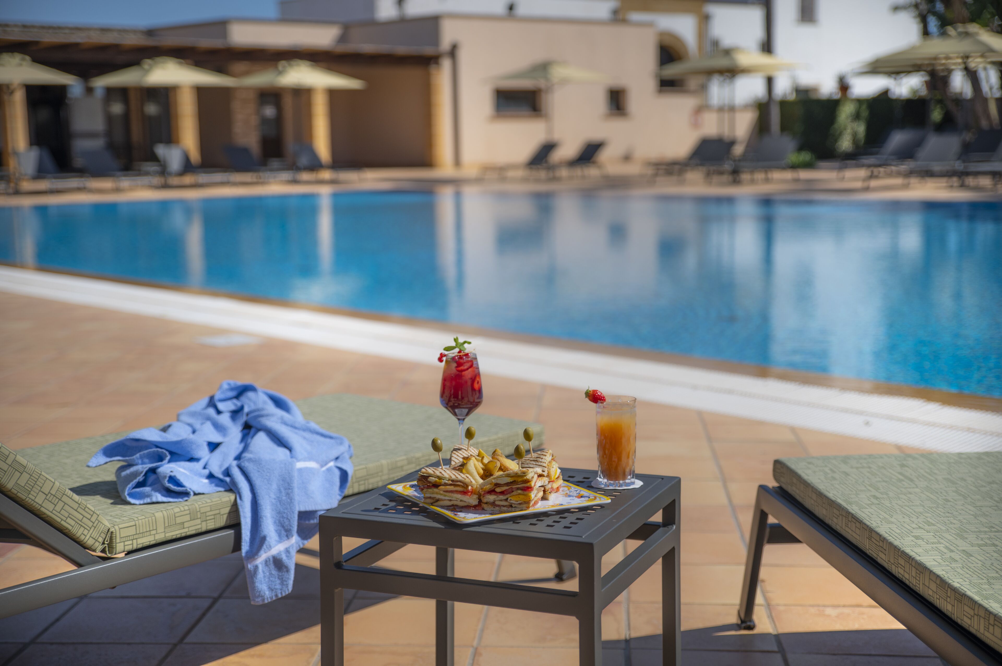Área da piscina com espreguiçadeiras, uma mesa com sanduíche e bebidas, e uma piscina ao fundo.