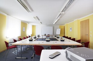 Uma sala de conferências com uma mesa comprida, cadeiras vermelhas, um quadro branco e um projetor.
