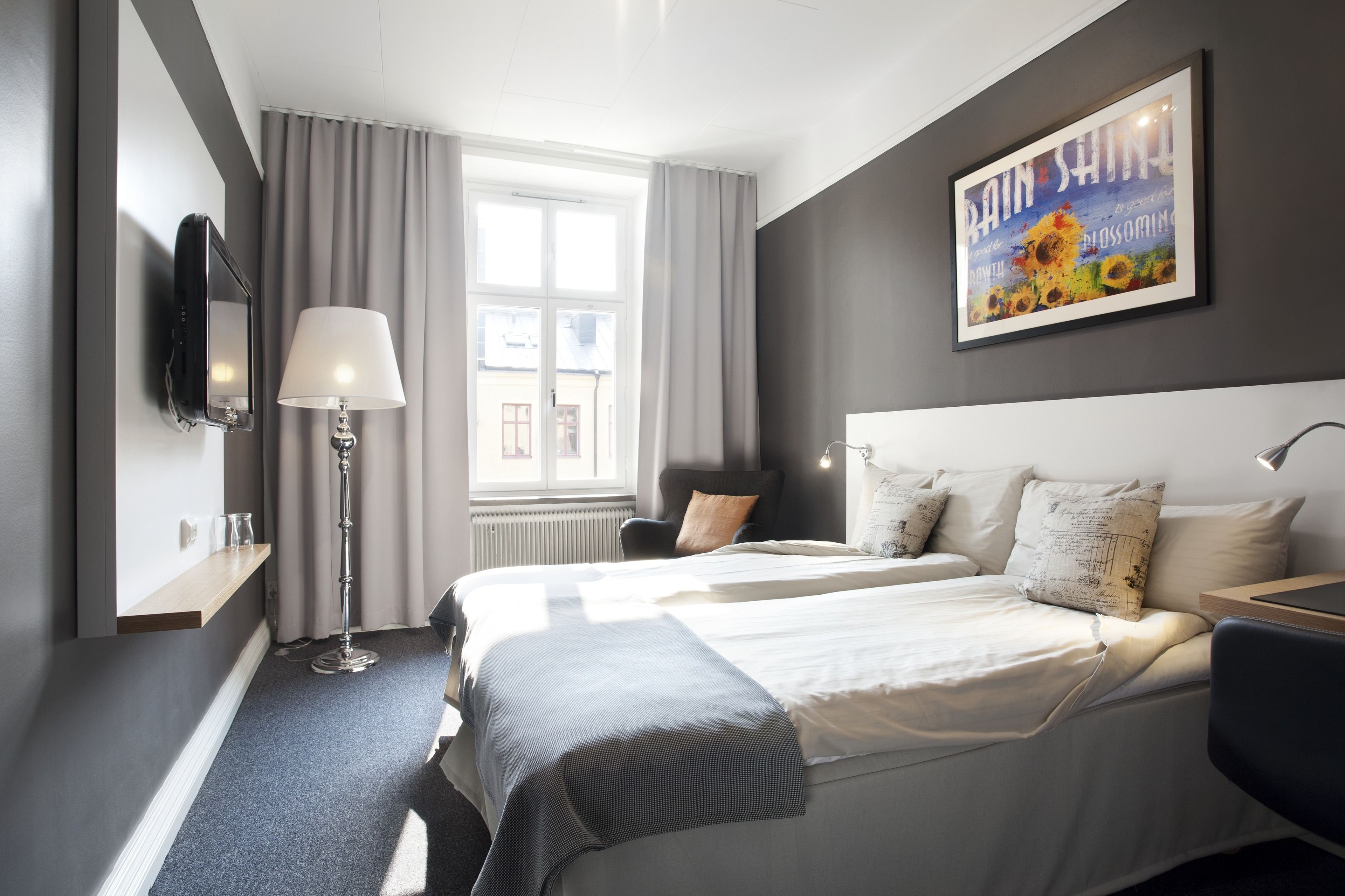 Quarto de hotel moderno com duas camas, uma janela grande com cortinas, um quadro na parede e um abajur.
