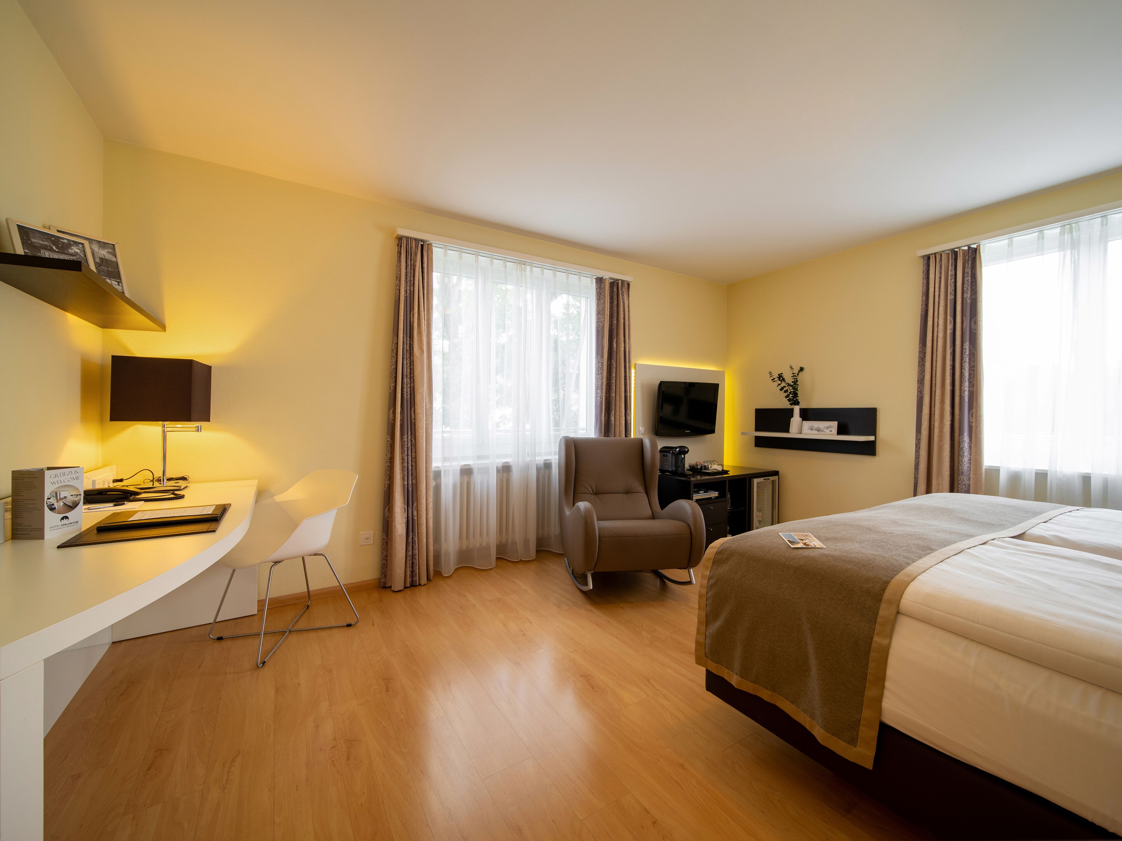 Quarto de hotel com cama, escrivaninha com luminária, cadeira, televisão e cortinas.