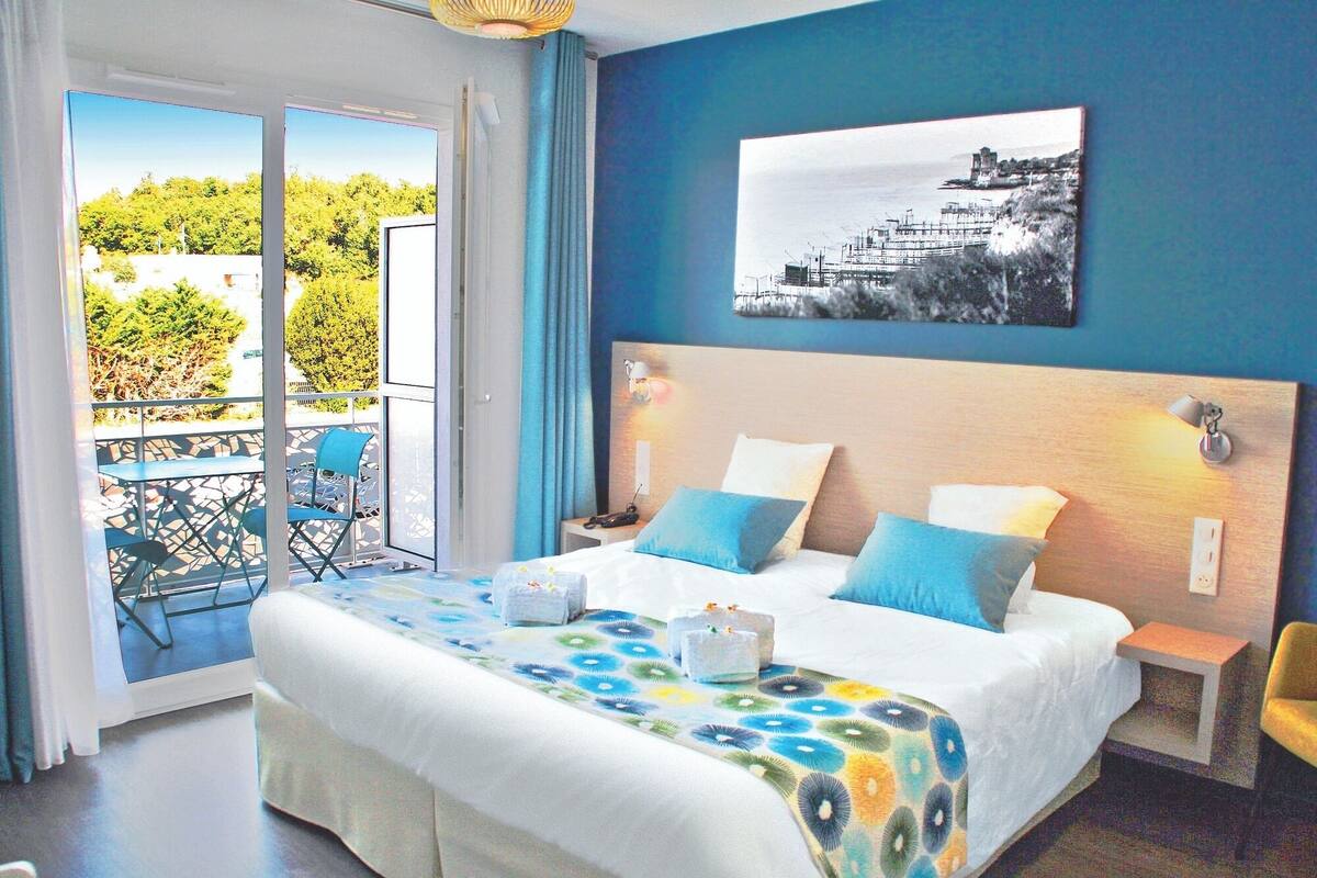 Quarto de hotel com cama, mesa de cabeceira, cadeira, varanda com mesa e cadeiras, e vista para áreas verdes e um edifício.