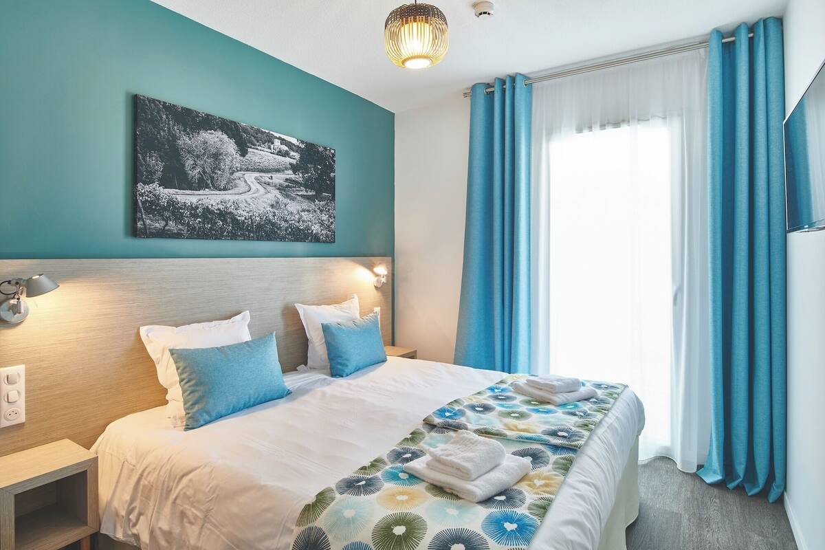 Quarto de hotel com cama, mesa de cabeceira, luminária na parede, um quadro e janela com cortinas.