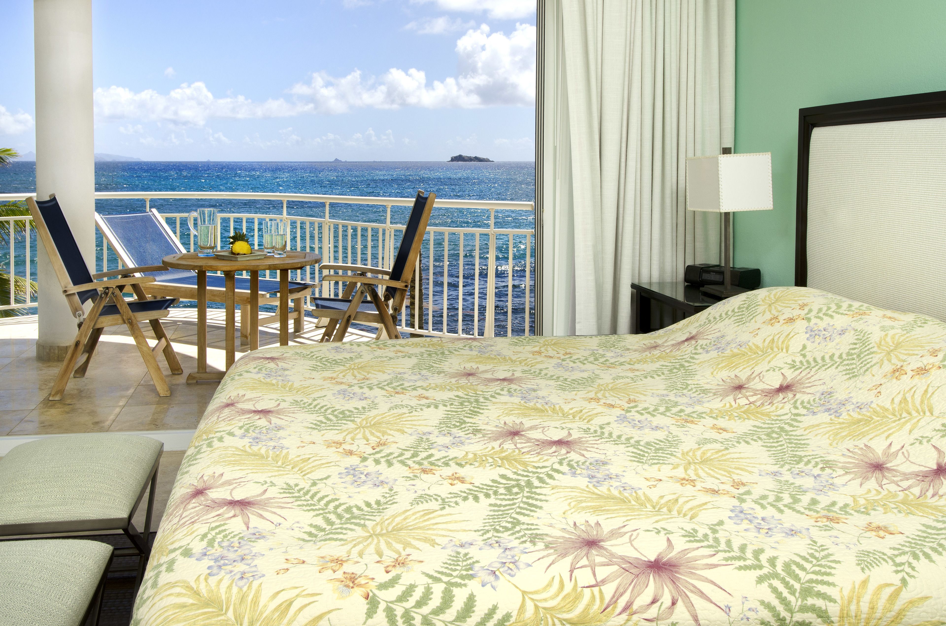 Quarto de hotel com varanda, cama com roupa de cama florida, mesa com cadeiras e vista para o mar.