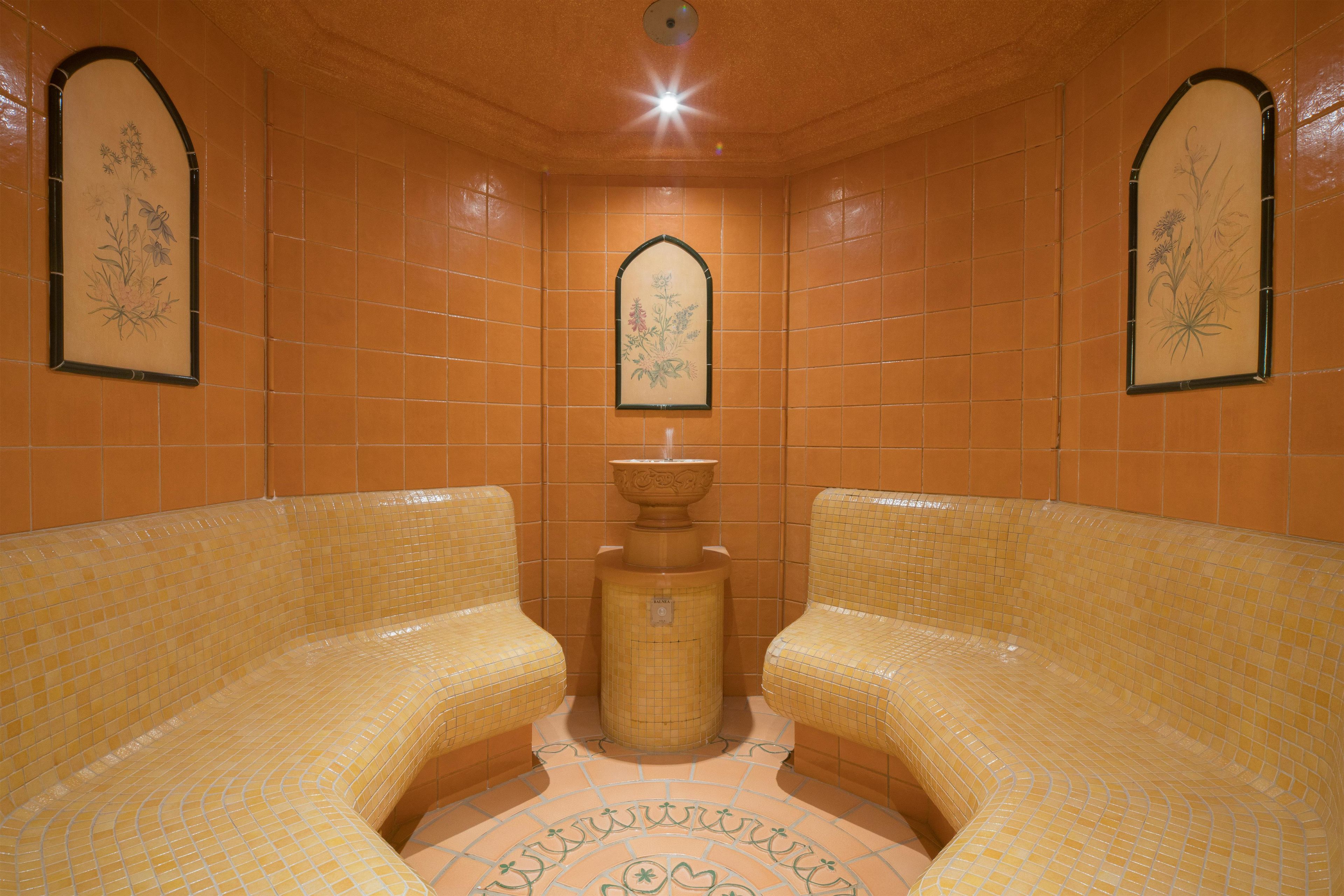 Um espaço de spa com duas áreas de assento acolchoadas, uma fonte central e azulejos decorativos nas paredes.