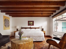 Quarto com uma cama grande, vigas de madeira, janela com vista, mesa de cabeceira com abajur e uma cadeira.