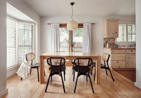 Uma área de jantar com uma mesa de madeira, quatro cadeiras, um lustre pendurado e vista para o exterior através de uma janela com cortinas brancas.