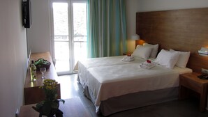 Quarto de hotel com uma cama grande, uma escrivaninha com computador, uma janela com cortinas e uma cabeceira de madeira.