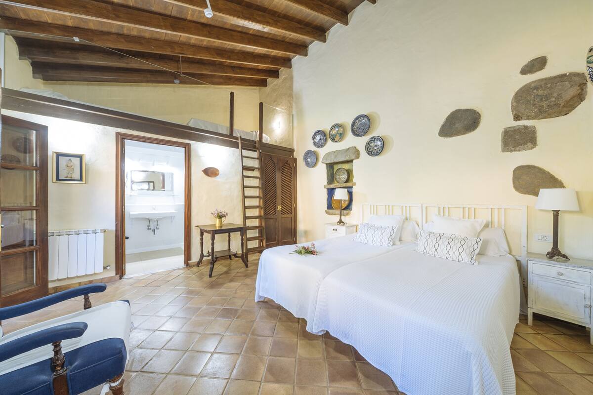 Quarto com cama grande, vigas de madeira no teto, banheiro visível por uma porta aberta e uma pequena mesa com um vaso.