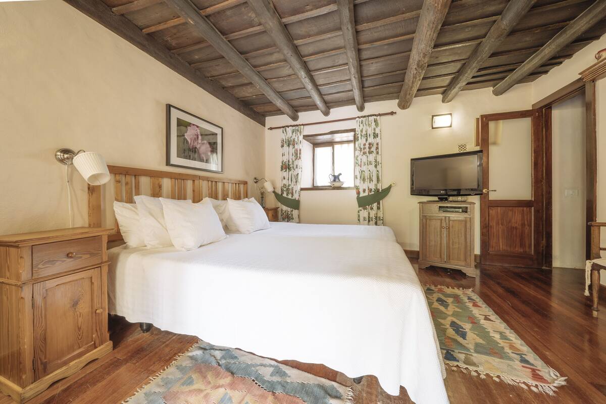 Quarto com cama de madeira, mesa de cabeceira, televisão e janela com cortinas.