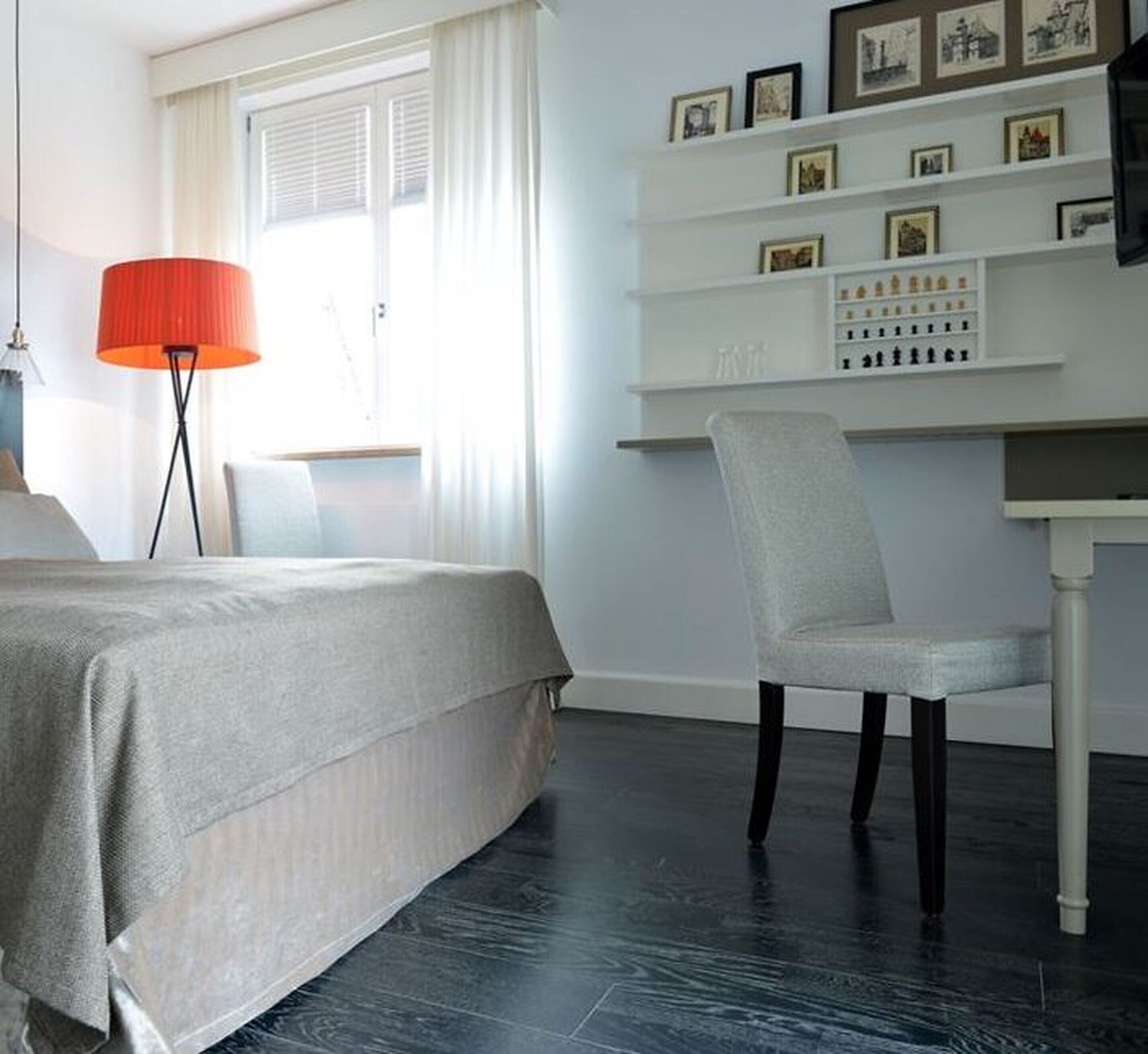 Quarto com cama, cadeira, escrivaninha e prateleira com quadros.