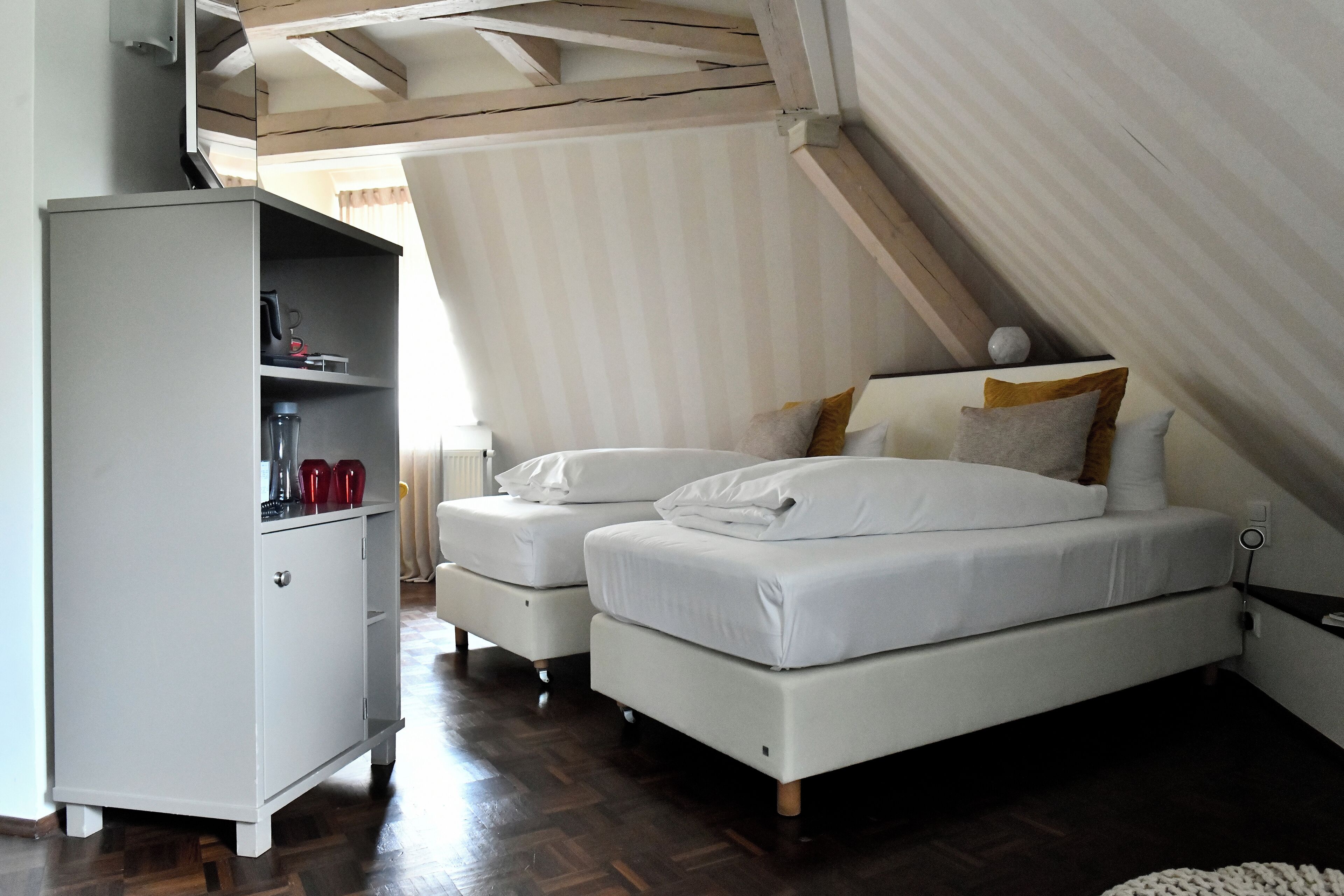 Quarto com teto estilo loft, cama branca com almofadas, guarda-roupa branco e pequena mesa com abajur.