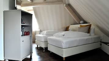 Quarto com teto estilo loft, cama branca com almofadas, guarda-roupa branco e pequena mesa com abajur.