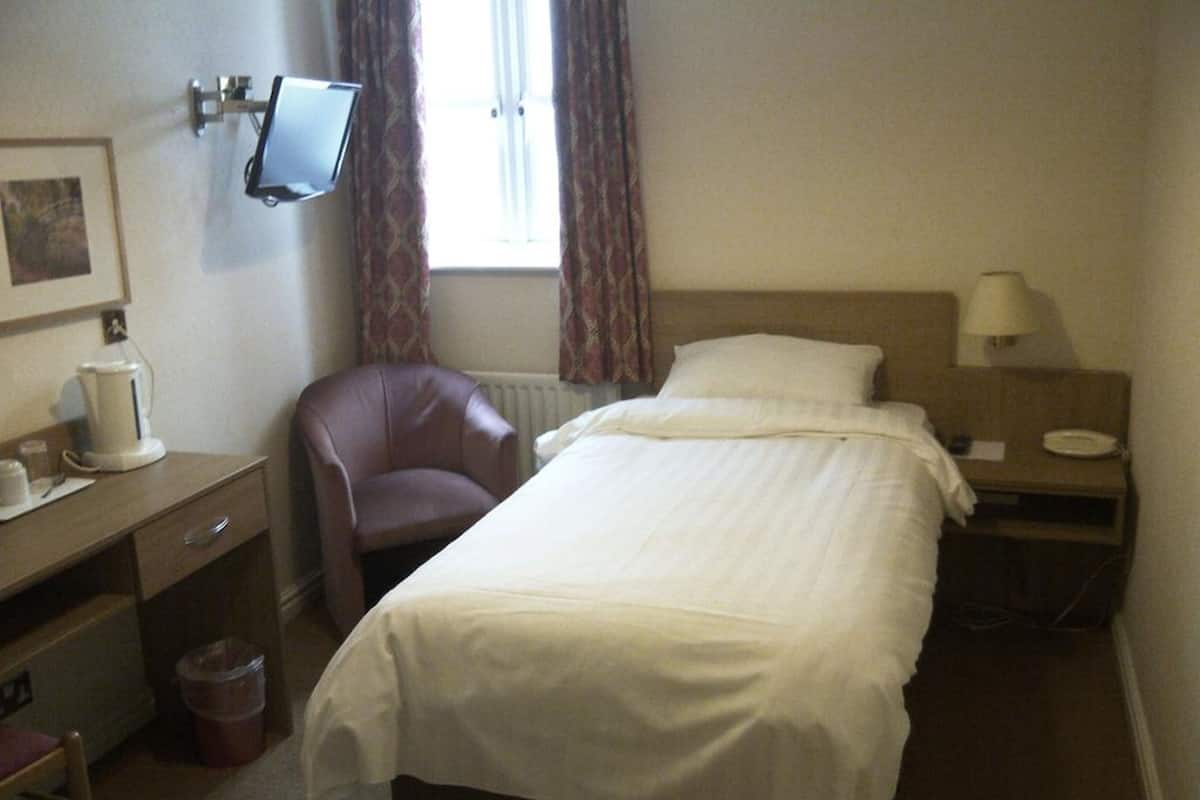 Quarto de hotel com uma cama, uma escrivaninha com monitor de computador, uma cadeira, uma mesa de cabeceira com abajur e uma janela com cortinas.