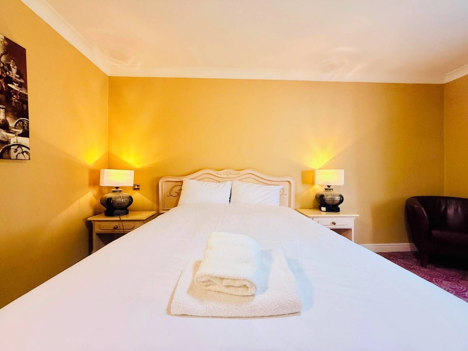 Quarto de hotel com uma cama grande, duas luminárias de cabeceira, uma cadeira e um quadro na parede.