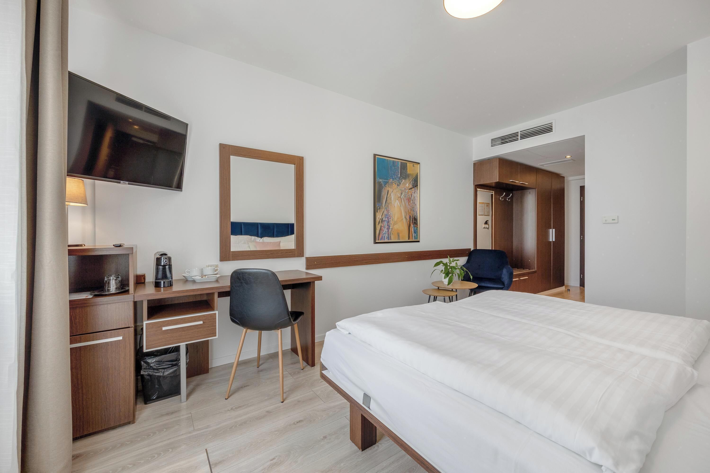 Quarto de hotel com uma cama, uma escrivaninha com cadeira, televisão, um quadro e um guarda-roupa.