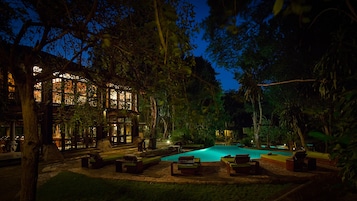 Vista noturna de uma área de piscina com um edifício com amplas janelas e uma piscina cercada por espreguiçadeiras e vegetação.