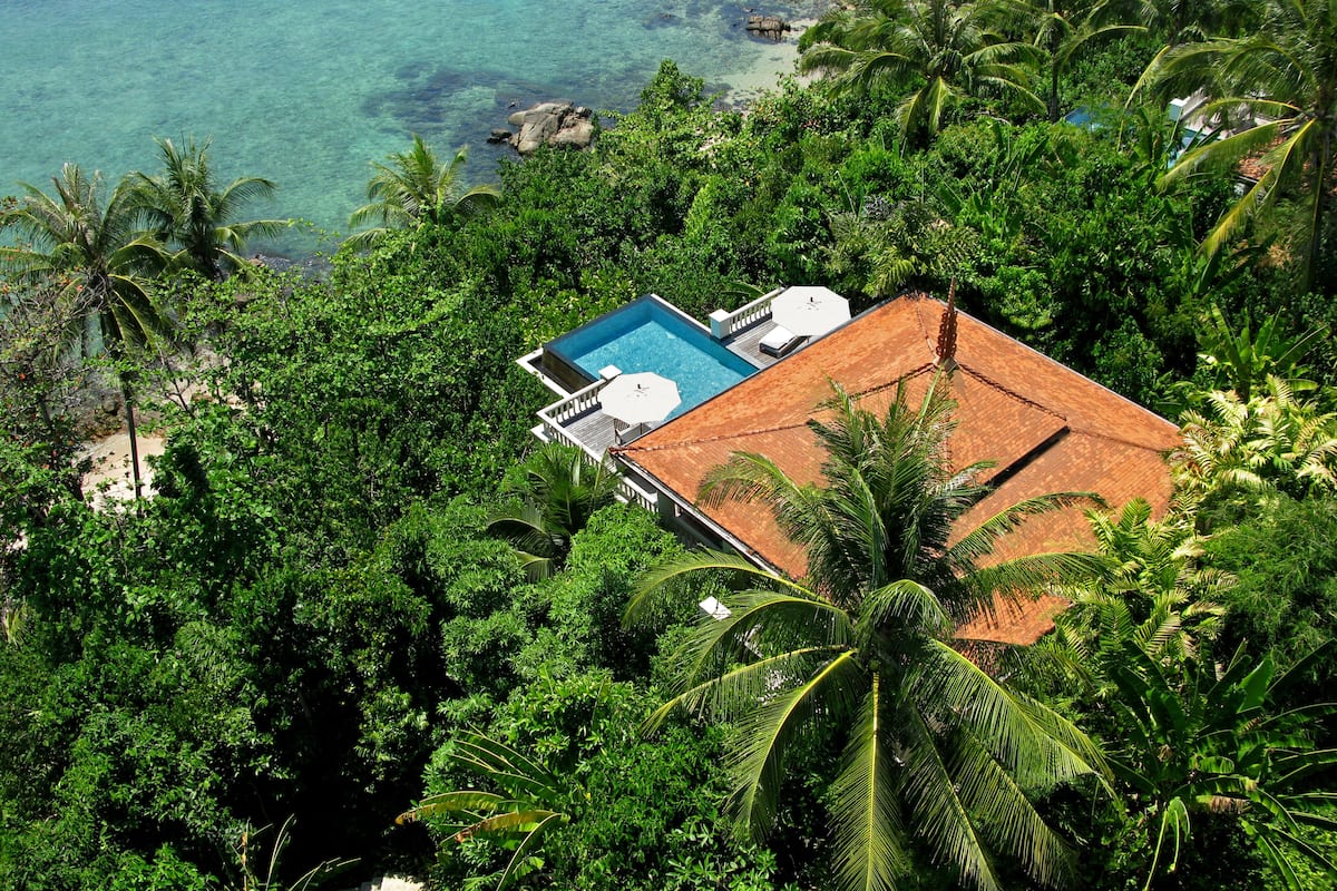 Vista aérea de um resort com piscina, cercado por vegetação exuberante e uma costa rochosa.