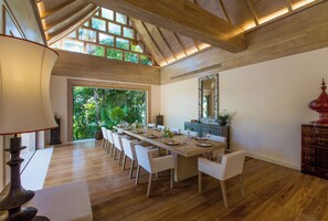 Uma sala de jantar com uma longa mesa de madeira posta para seis pessoas, cadeiras brancas, um lustre e uma janela grande com vista para o verde.