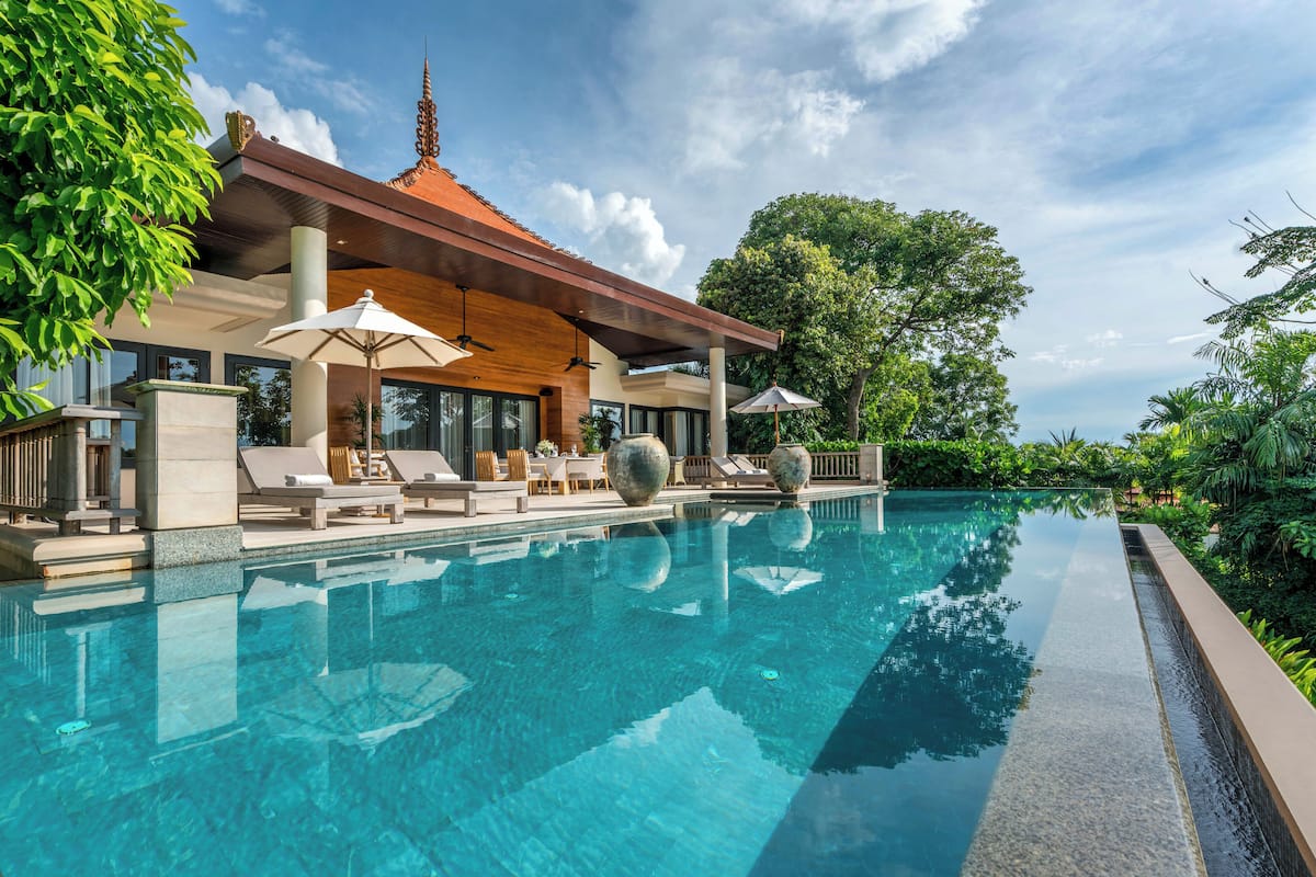 Piscina com espreguiçadeiras e guarda-sóis, rodeada por vegetação exuberante e um edifício com telhado de azulejos.