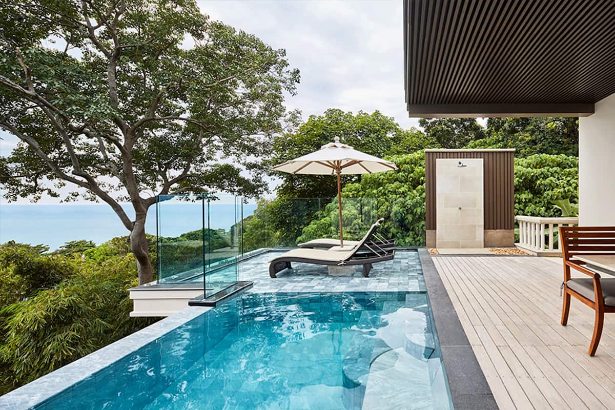Uma piscina infinita com borda de vidro, uma espreguiçadeira e um deck de madeira.