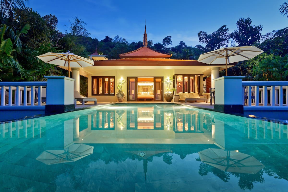 Vista da piscina de um resort, com um edifício bem iluminado, piscina cristalina e vegetação exuberante ao fundo.