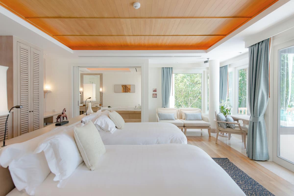 Quarto de hotel moderno com duas camas grandes, teto de madeira, área de estar com sofá e cadeiras, e janelas amplas com cortinas.