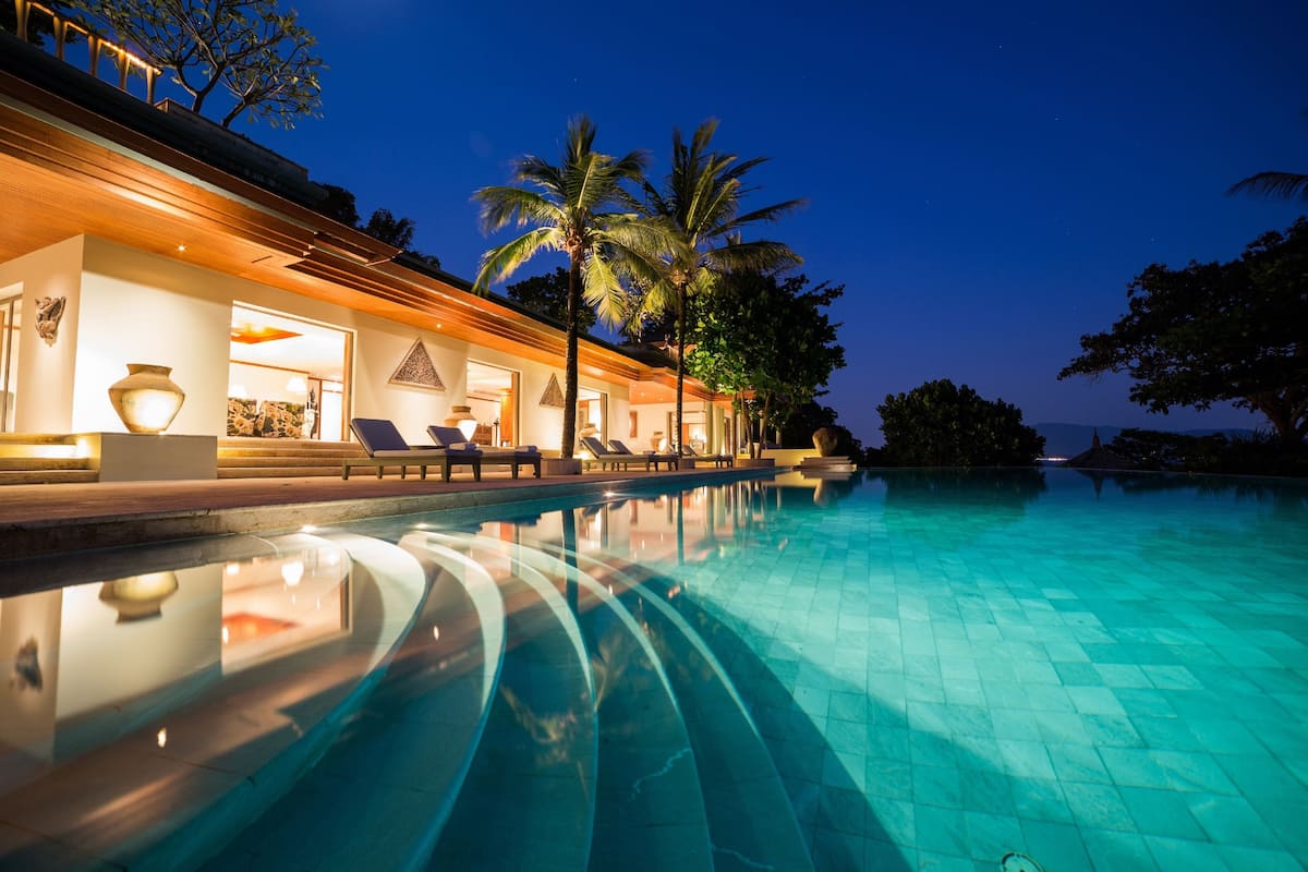 Uma área na piscina com espreguiçadeiras, um edifício bem iluminado e palmeiras.