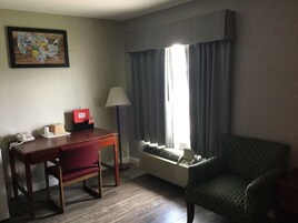 Quarto de hotel com escrivaninha, cadeira, abajur e janela com cortinas.