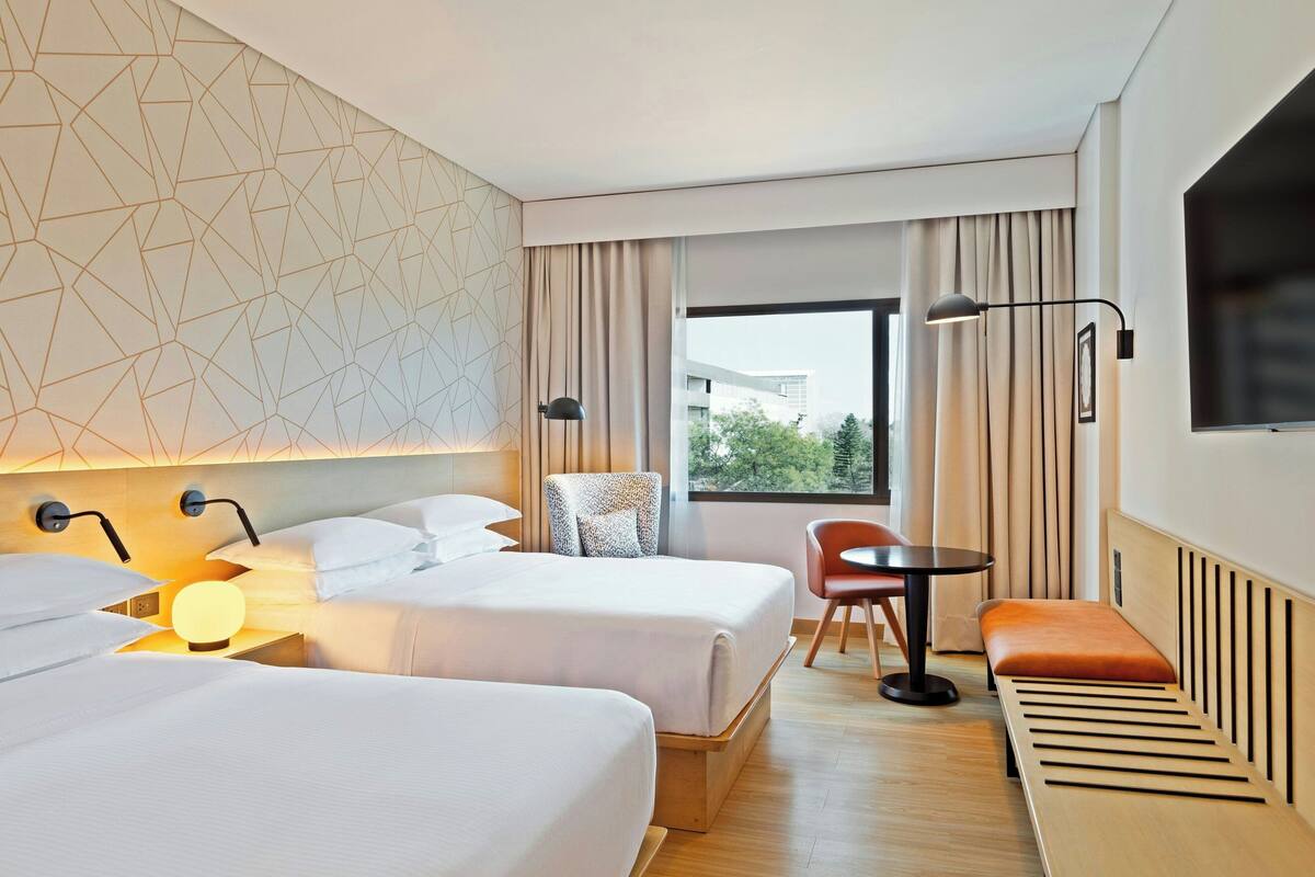 Quarto de hotel com duas camas, área de estar, televisão e janela com vista para edifícios.