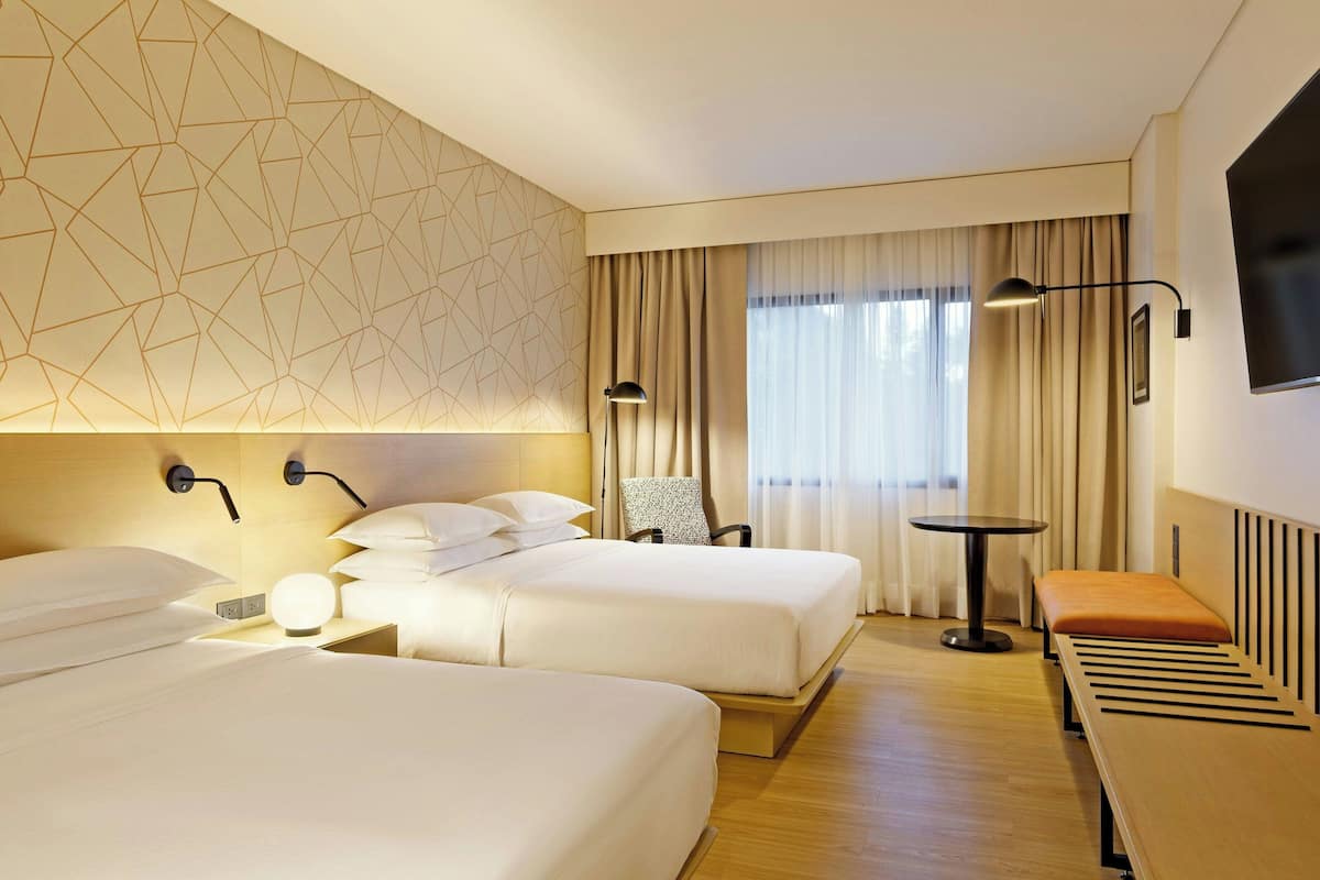 Quarto de hotel com duas camas, uma mesinha, um banco, uma cadeira e uma televisão na parede.