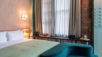 Quarto de hotel com cama, mesa de cabeceira com abajur, janela com cortinas e parede de tijolos.
