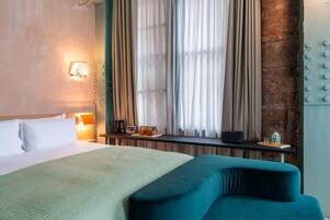Quarto de hotel com cama, mesa de cabeceira com abajur, janela com cortinas e parede de tijolos.