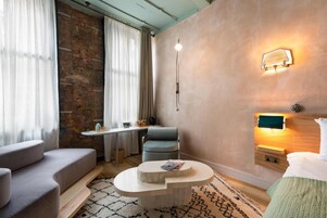 Quarto de hotel moderno com parede de tijolos, mesa de centro redonda, sofá, mesinha com abajur e cama com manta verde.
