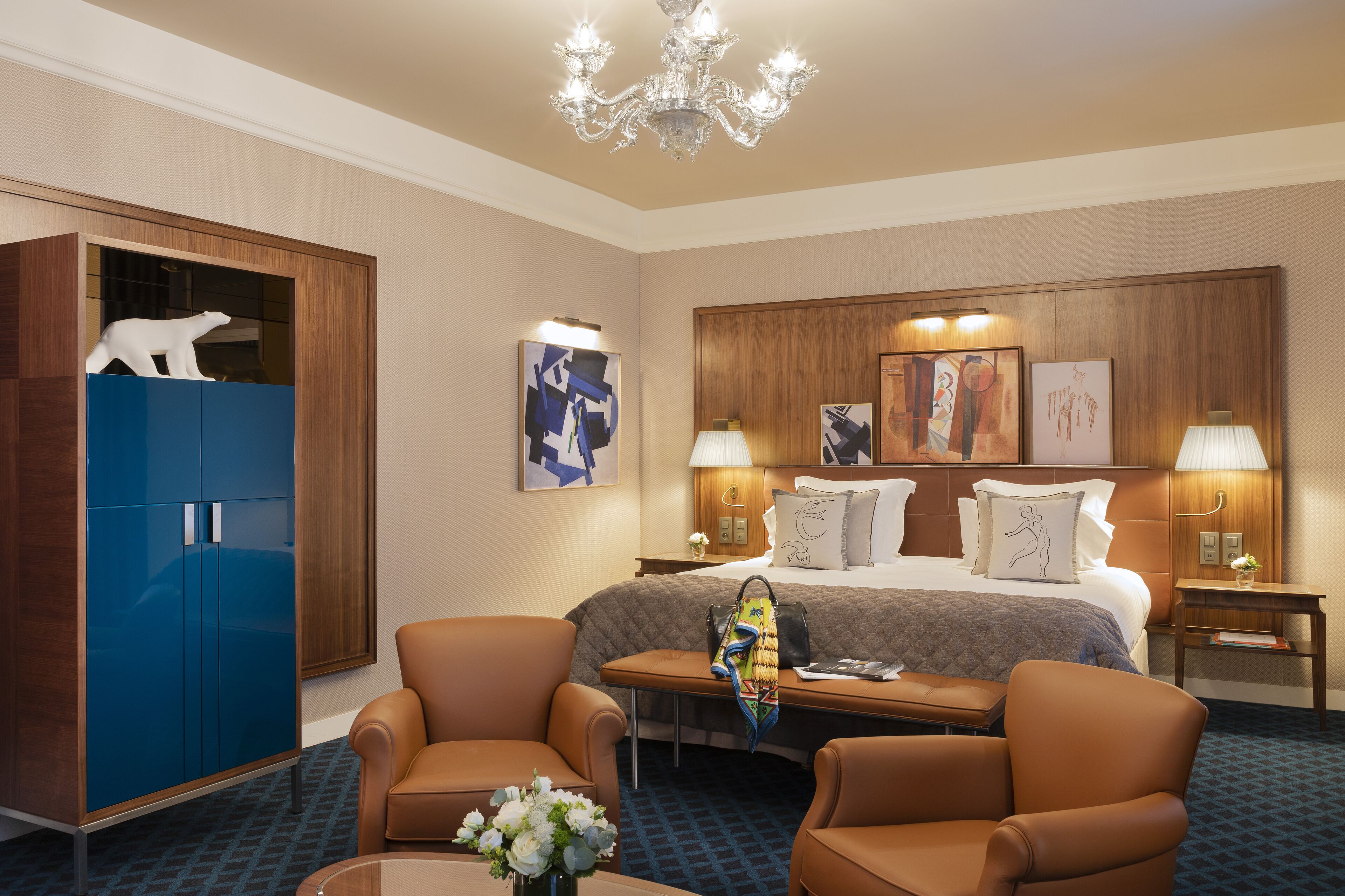 Quarto de hotel com uma cama grande, duas luminárias laterais, uma cabeceira de madeira, um guarda-roupa com uma figura de urso polar e um candelabro.