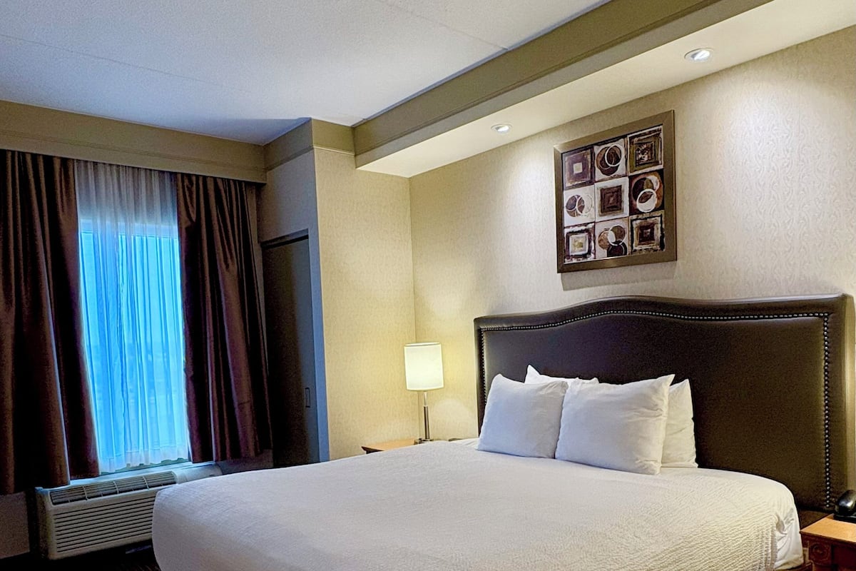 Quarto de hotel com uma cama grande, mesa de cabeceira, abajur, relógio de parede e janela com cortinas.