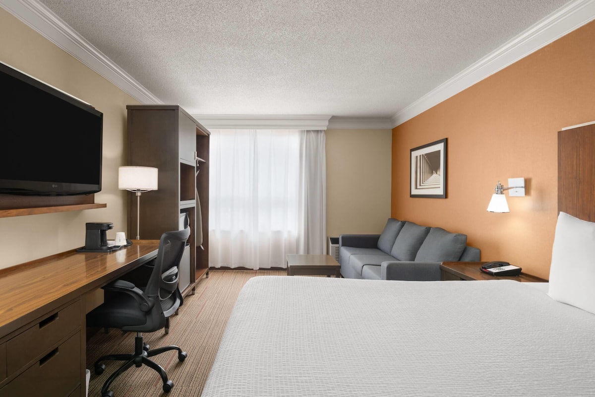 Quarto de hotel com cama, escrivaninha com cadeira, televisão, sofá e janela com cortinas.