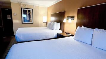 Quarto de hotel com duas camas, um quadro pendurado na parede, mesa de cabeceira com abajur e uma porta.