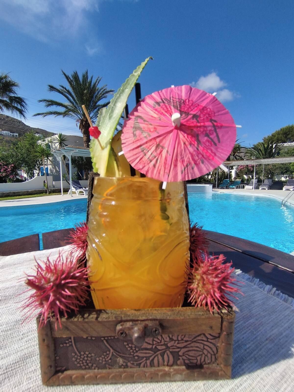 Disposição de bebidas tropicais com um guarda-sol rosa, um vaso decorativo e uma caixa de madeira com alça, colocada à beira da piscina, com espreguiçadeiras e palmeiras ao fundo.