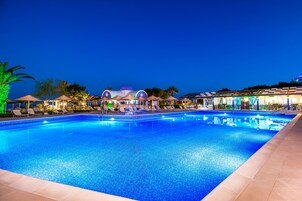 Piscina com espreguiçadeiras e guarda-sóis, cercada por palmeiras e um edifício com telhado em cúpula.