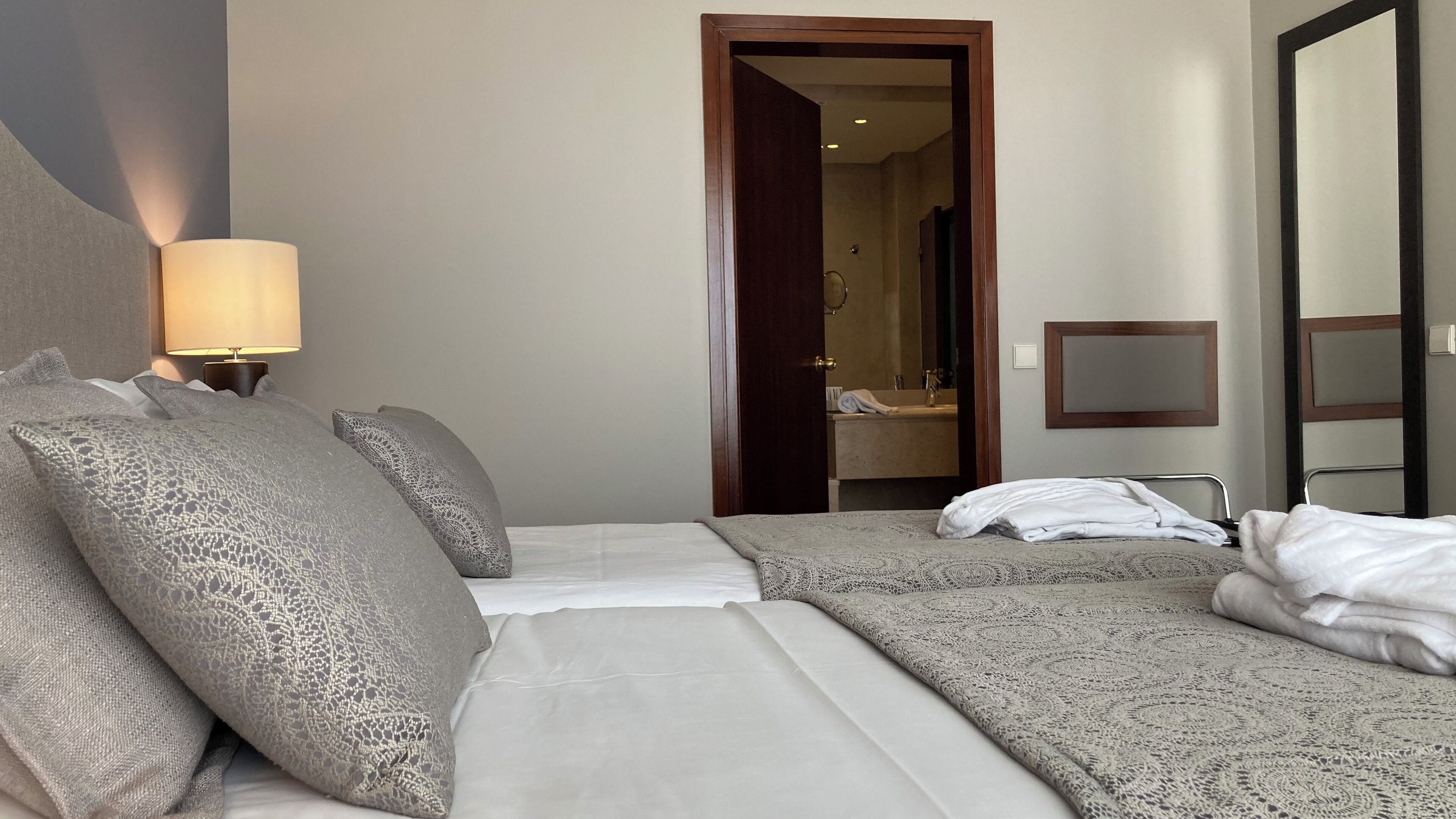 Quarto de hotel com duas camas, uma mesa de cabeceira com abajur, banheiro visível através de uma porta aberta e um espelho grande na parede.